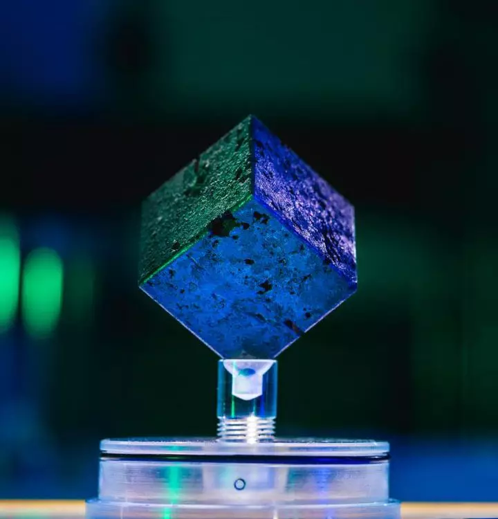 Una imagen azul y verde de un pequeño cubo de metal que descansa sobre una pantalla, con luces de colores en el fondo