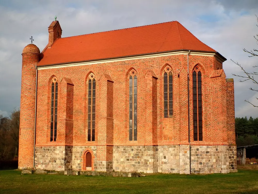 An taobh amuigh de shéipéal brící oráiste le fuinneoga caola, arda Gotach, díon beannach dearg agus corp cuartha ar thaobh amháin.