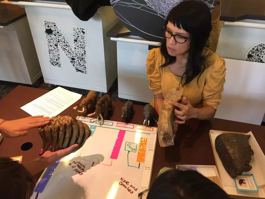 Женщина в очках и с темными волосами сидит за деревянным столом, держа в руках 3D-сканированное окаменелость, и рассказывает посетителям музея об окаменелостях и науке.