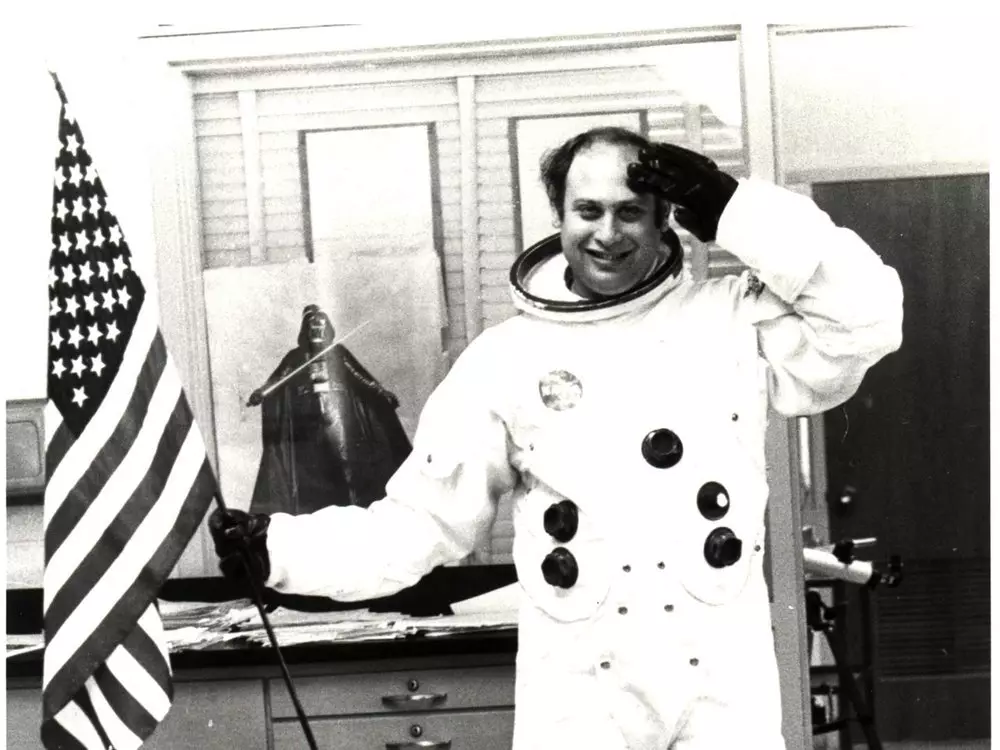 Retrat informal de l'aula del professor Herbert Stephen Desind (1945-1992), vestit amb un vestit espacial de reproducció de l'era Apol·lo, sostenint una bandera americana; cap a la dècada de 1980. Desind era un aficionat als vols espacials, i la seva col·lecció de fotografies d'avions i naus espacials va ser donada al Museu Nacional de l'Aire i l'Espai l'any 1997. Aquesta imatge forma part de la col·lecció Herbert Stephen Desind.