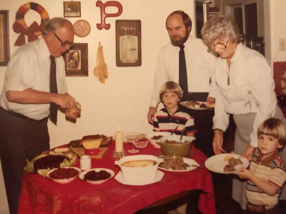 Drie volwassenes staan en twee klein kindertjies staan om 'n tafel met bord. Die tafel word gedek deur 'n rooi tafeldoek en het 'n groot kers in die middel met kos rondom gerangskik.