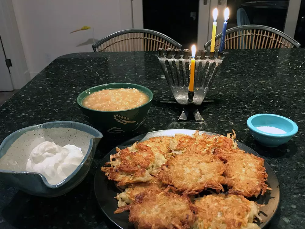 Un piatto pieno di frittelle di patate dorate è posto su un tavolo da pranzo. Dietro di loro ci sono ciotole di panna acida, salsa di mele, sale e una menorah con tre candele accese.