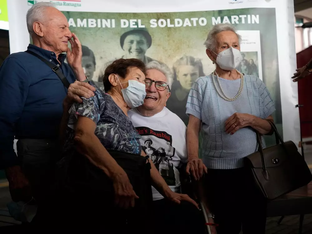 Adler stă într-un scaun cu rotile, în centru, îmbrățișând-o pe Mafalda, în timp ce Bruno și Giuliana zâmbesc și pozează pe ambele părți.