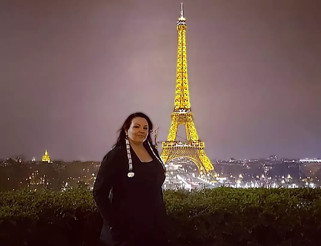 La dissenyadora de moda Norma Baker–Flying Horse, vestida amb un vestit negre i guants, es troba a l'aire lliure a París de nit. La Torre Eiffel està il·luminada al fons.