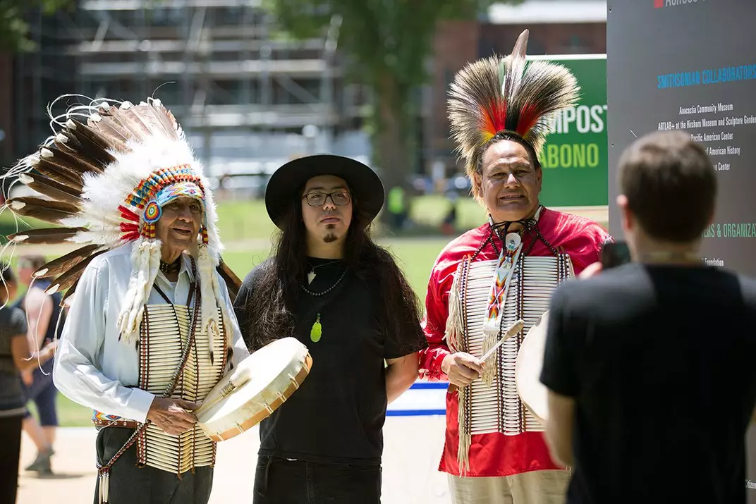 Tre mænd står sammen og smiler. Mændene til venstre og højre er iført traditionelle fjerbeklædte hovedbeklædninger, og manden i midten har langt hår og bærer en sort, bredskygget hat.