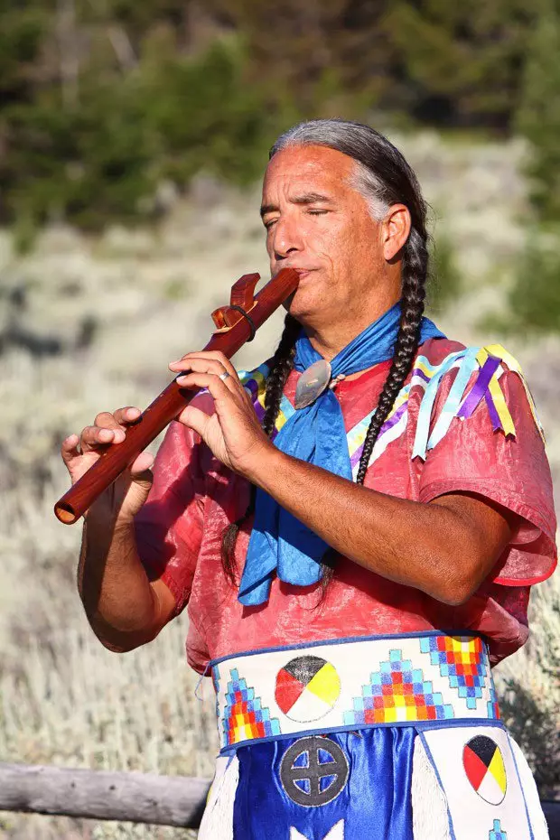 Un hombre con largas trenzas y atuendos rosas, azules y blancos toca una larga flauta de madera.