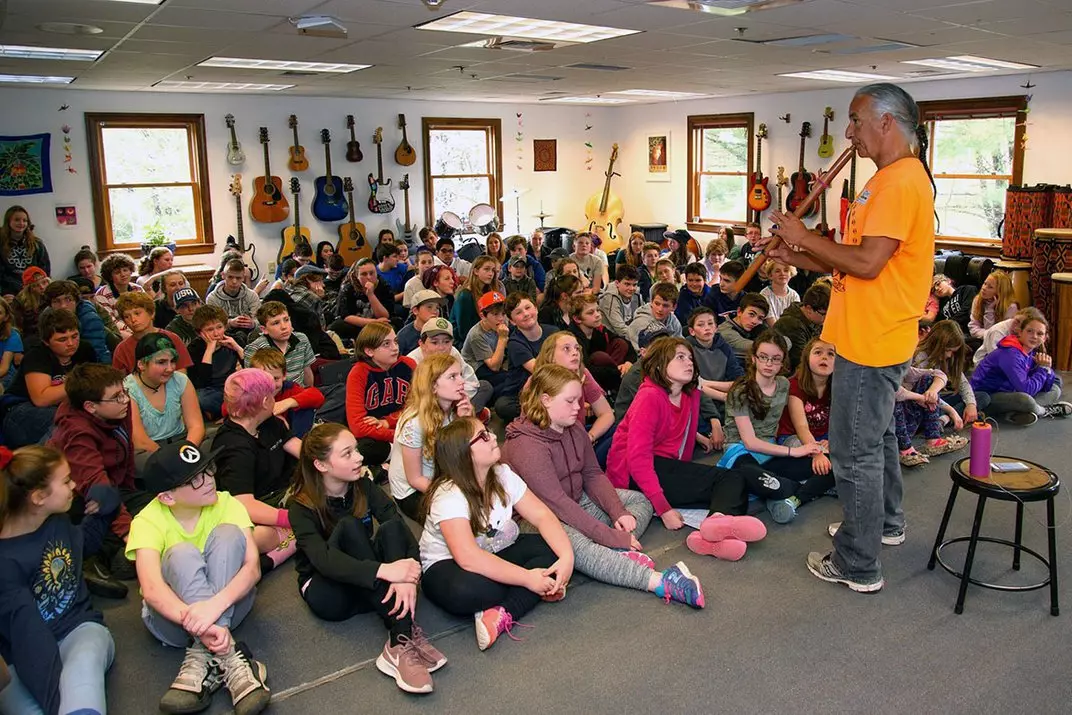 Man met lange vlechten en felgeel shirt speelt fluit voor een vol klaslokaal met kinderen die op de grond zitten.
