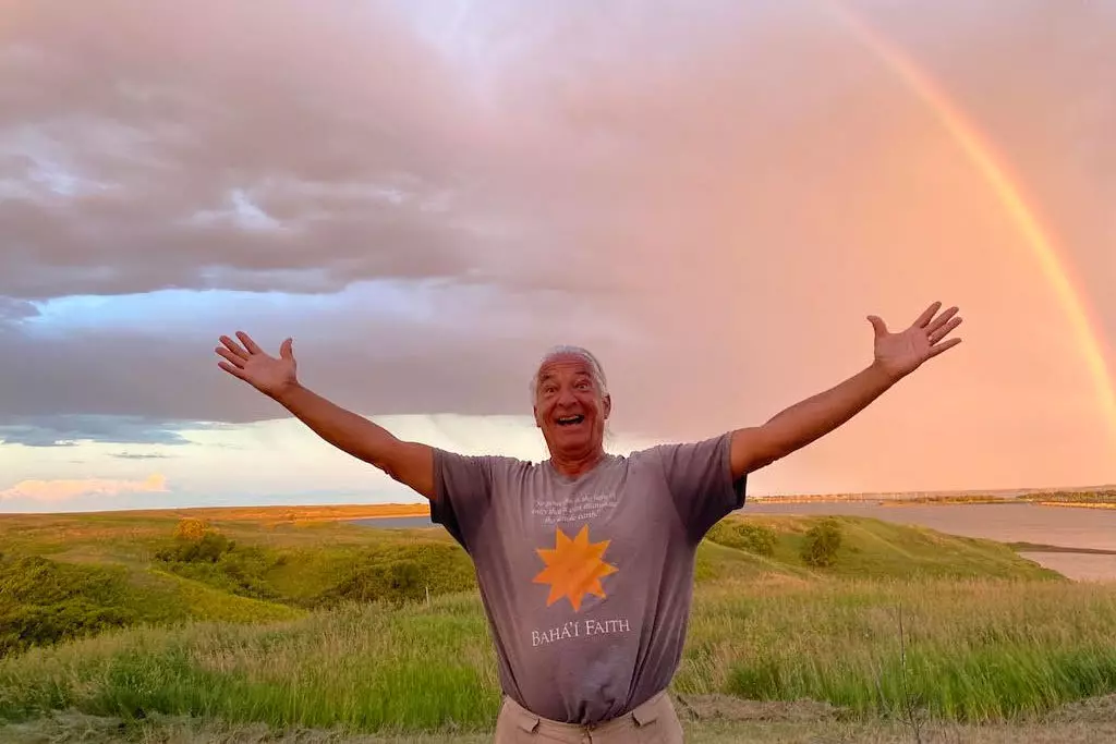 El hombre posa al aire libre, con los brazos extendidos, como si mostrara el arcoíris que se extiende sobre su cabeza.