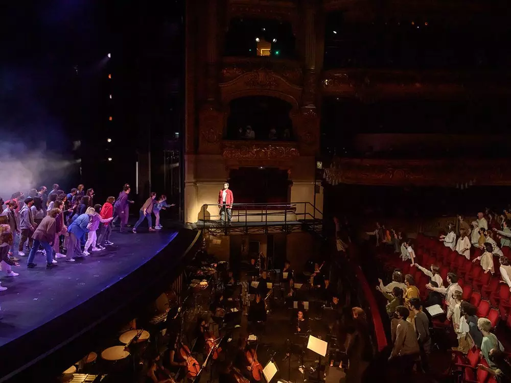 Uma apresentação em uma sala de ópera, olhando de lado para o fosso da orquestra. Os artistas estão tanto no palco, iluminados em azul à esquerda, quanto nas primeiras fileiras de assentos vermelhos, à direita.