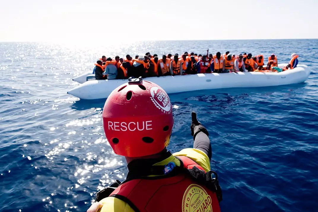 Z łodzi osoba w ekwipunku ochronnym i czerwonym kasku z napisem RESCUE wskazuje na nadmuchiwaną tratwę pełną kilkudziesięciu osób w pomarańczowych kamizelkach ratunkowych.