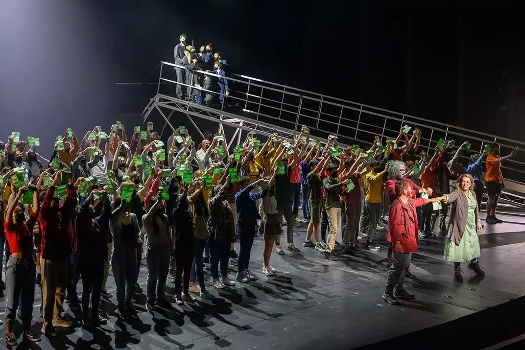 Decenas de intérpretes no escenario, todos menos un par diante sostindo papeliños verdes. Unha rampla no escenario suxire a posta en escena dun barco que chega a un peirao.