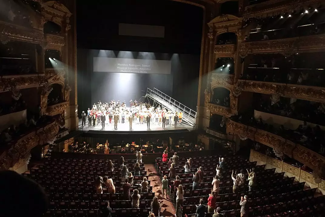 Els membres dispersos d'un públic fan una ovació de peu als intèrprets fent reverències a l'escenari d'una sala d'òpera.