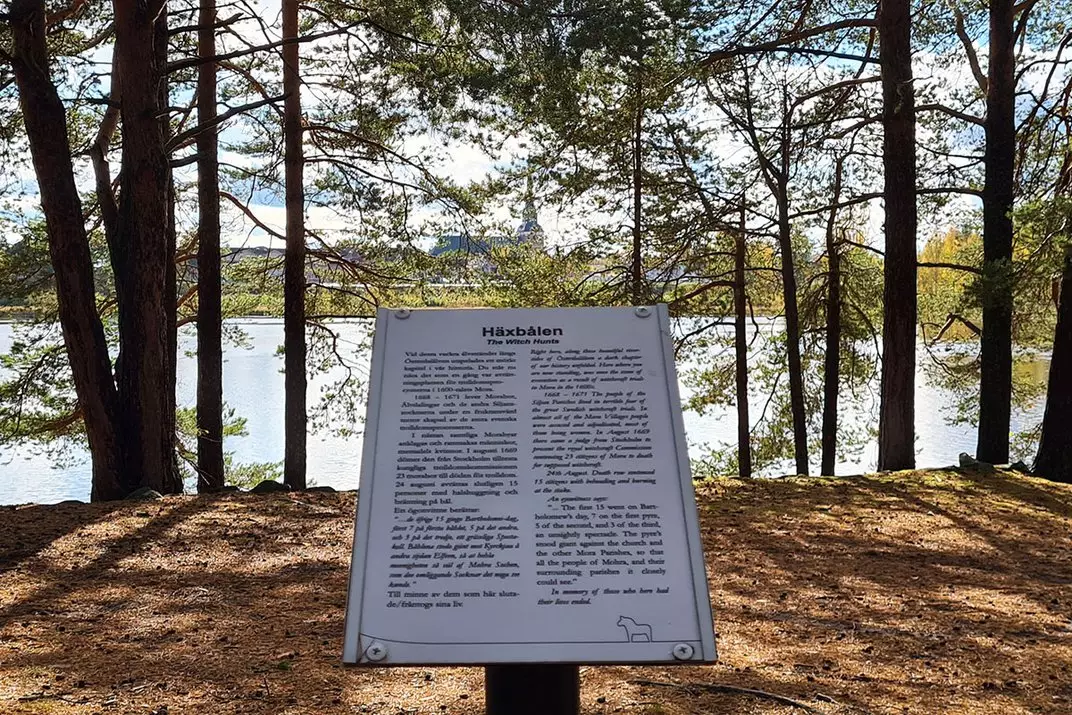 Komemorativni znak, napisan na švedskom, na naizgled idiličnom prizoru drveća uz rub rijeke.