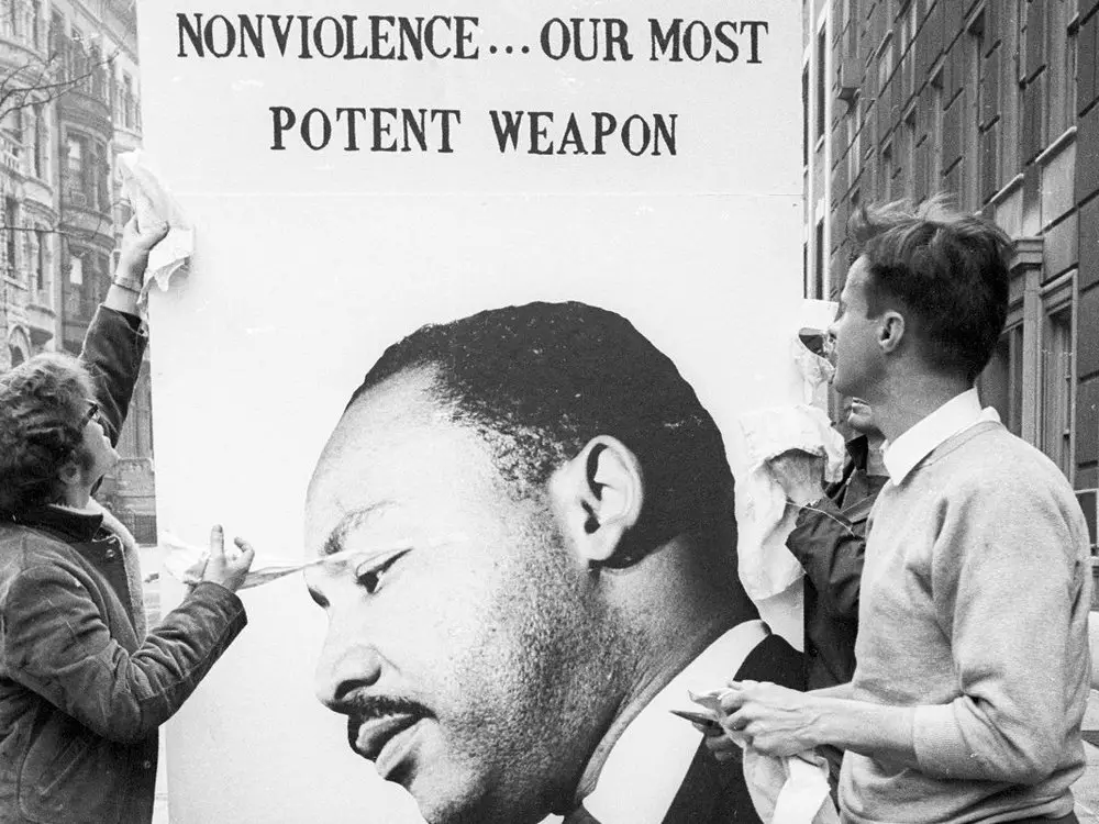 Doi civili se uită la un afiș care prezintă chipul lui Martin Luther King Jr., capul acestuia înfățișat sub cuvintele „Nonviolență... cea mai puternică armă a noastră”.
