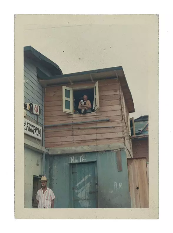 Снимка на дървена къща, боядисана в синьо с червени капаци. Една жена се навежда от прозореца на горния етаж и държи куче, а мъж е долу на улицата и пуши цигара.
