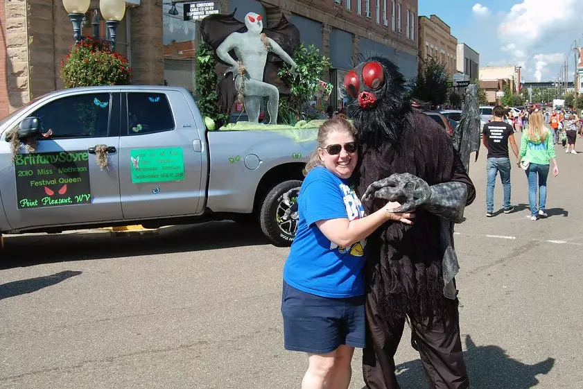 Op engem Outdoor Street Festival, eng Fra ëmfaasst eng Persoun eis an engem donkel fuzzy Mothman Kostüm gekleet, mat enger donkel Mask mat roude Aen a Mond.