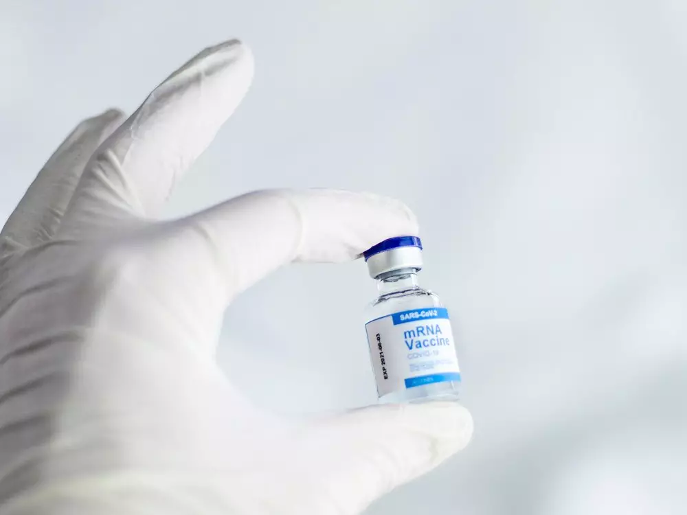 Lijeva ruka u bijeloj rukavici drži malu staklenu bočicu koja sadrži doze pfizer cjepiva covid-19.
