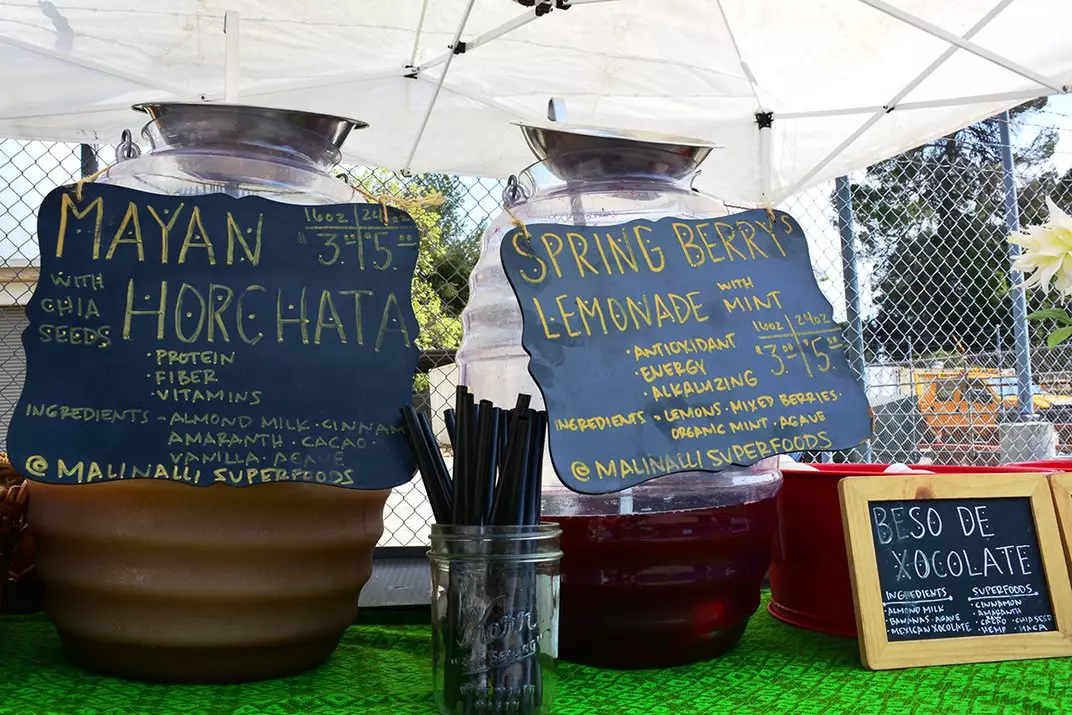 Tvertnes, kas piepildītas ar svaigiem, mājās gatavotiem dzērieniem, ir novietotas uz galda. Uz konteineriem ir piekārtas ar rokām apgleznotas zīmes, kas apraksta katru dzērienu: Mayan Horchata un Spring Berry Limonade.