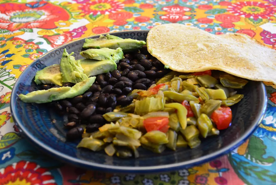 Čerstvo uvarený tanier s fazuľou, avokádom, restovanou zeleninou a kukuričnou tortillou sa položí na svetlý, kvetinový obrus.