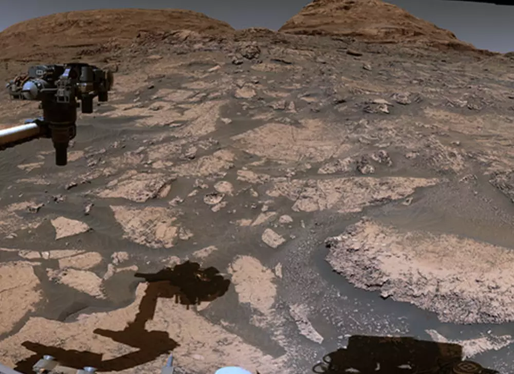 Curiosity Mars Roverilla otettu kuva. Kuvassa näkyi Marsin pölyinen pinta ja muutama kukkula taustalla. Kuvassa näkyy myös roverin käsi.