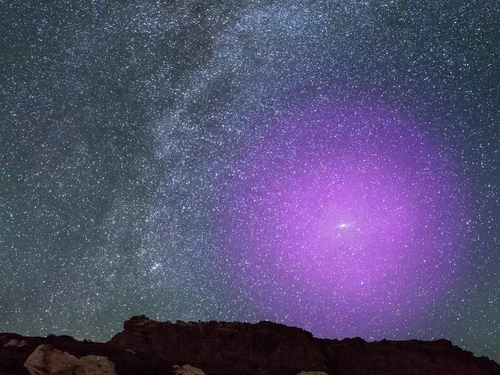 Вид на скелі на передньому плані з великим блискучим нічним небом позаду - з'являється багато зірок і крива Чумацького Шляху; у нижній правій частині неба ілюстроване велике коло фіолетового світла вказує, де буде ореол Андромеди