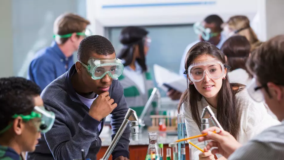 Mladí dospelí študenti rôzneho etnického pôvodu robia v triede chemický experiment. Všetci žiaci majú nasadené ochranné okuliare.