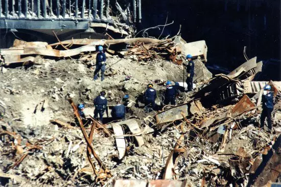 Foto van die vernietiging na 11 September 2001 in New York Stad. Werkers in blou helms en uniforms skarrel om 'n hoop verwoesting, gebuigde metaal en ruïnes.