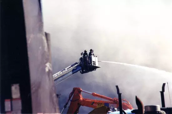 Pasaulio prekybos centro svetainės nuotrauka po 2001 m. rugsėjo 11 d. išpuolių. Du ar trys darbininkai su kietomis skrybėlėmis ir uniformomis yra krano rinktuve ir šaudo gaisrine žarna žemyn švelnia arka. Kairė matosi apdaužyta siena arba pastato dalis.