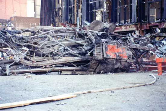 Foto van verwoesting in New York City na 11 September 2001. Ineengestorte deel van 'n gebou wat 'n industriële voorkoms met baie pype, kolomme en drade. In agtergrond, stukkende vensters. As en grys.