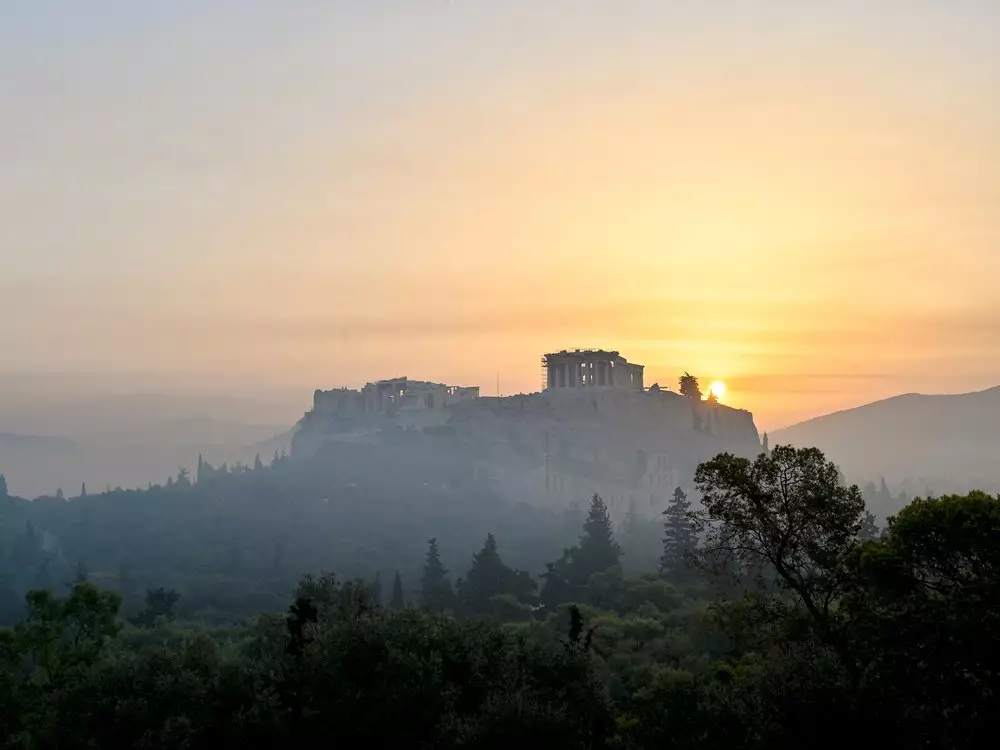 En vy av Parthenon i fjärran på en kulle, med solen lågt på himlen bakom ett töcken av rök