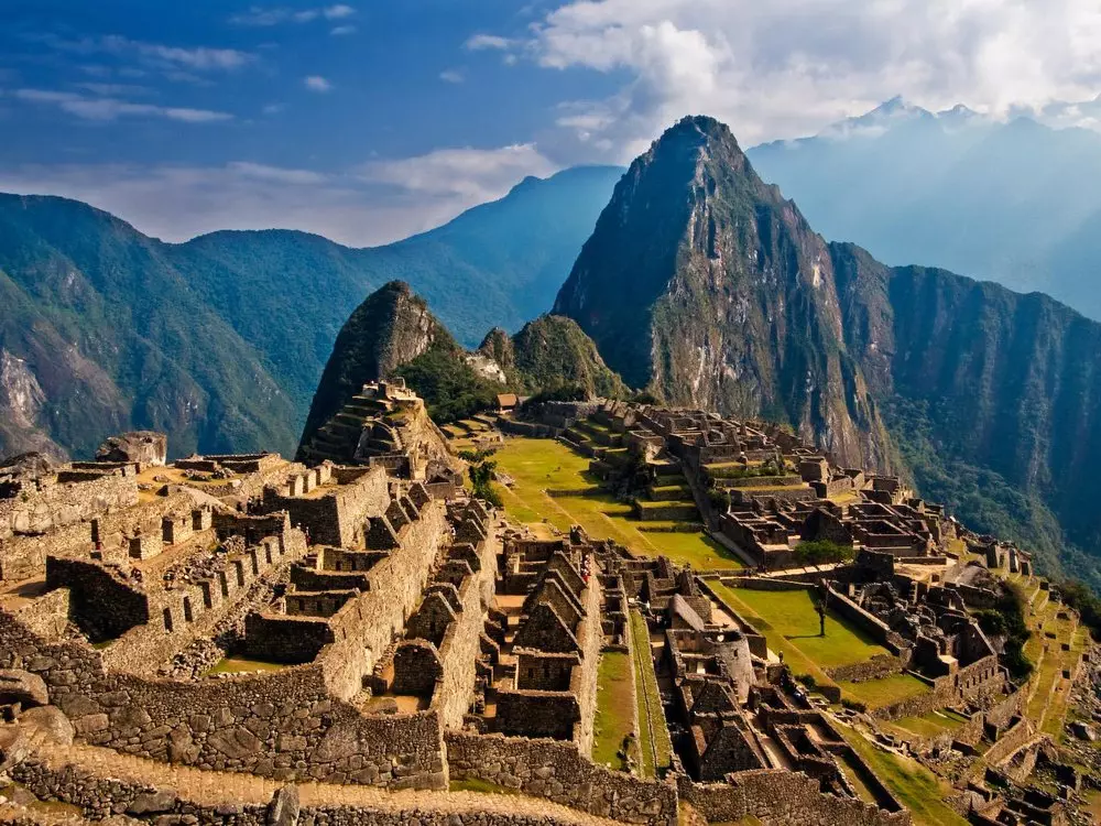 Eine Luftaufnahme von Machu Picchu bei Sonnenaufgang, mit einem orangefarbenen Licht, das die Steinstrukturen aus dem 15. Jahrhundert färbt, und einem weit entfernten Gipfel, der mit grünem Laub bedeckt ist