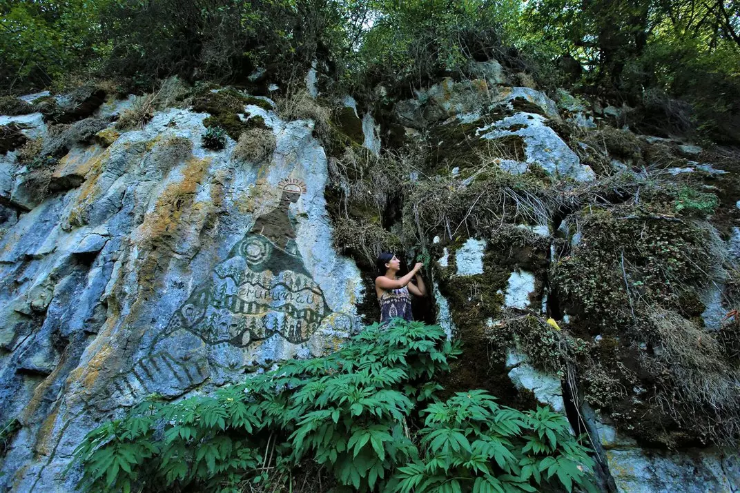 Afukidzwa nematombo ane mossy, mumwe mukadzi wechidiki akabata chiratidzo anomira pamberi pemadziro ematombo akavezwa nepetroglyphs.