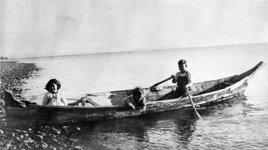 세 명의 어린 아이들이 바위가 많은 해안가에 정박해 있는 카누에 앉아 있습니다. 한 아이가 물에 두 개의 노를 들고 있습니다. 흑백 아카이브 사진입니다.