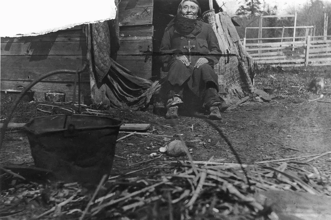 Un home está sentado na porta dunha cabana de madeira. En primeiro plano hai un fogar ao aire libre e un pote negro. Fotografía de arquivo en branco e negro.