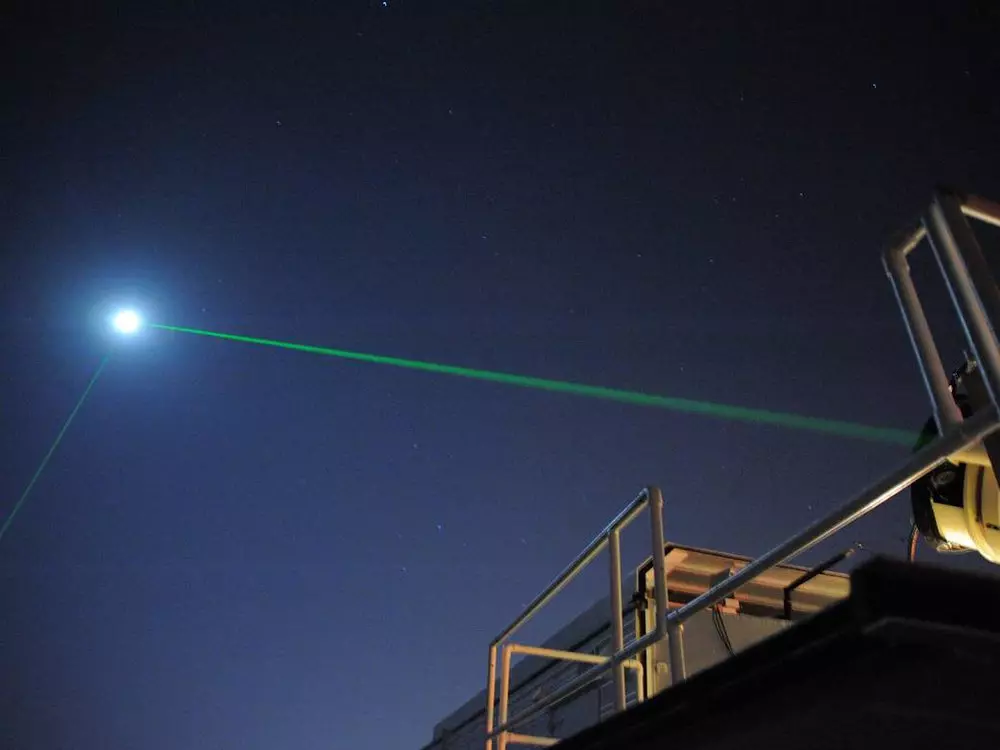 A dark night sky with some metal equipment and beams in the foreground, and then a bright green line shooting through the sky toward a bright full white moon