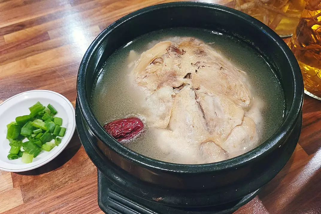 نمای نزدیک از یک کاسه سوپ با یک تکه بزرگ مرغ، در کنار یک ظرف کوچک از پیاز سبز خرد شده.