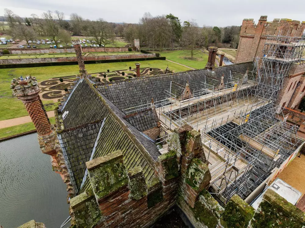 Une vue plongeante sur le toit du Manoir, avec environ la moitié de son toit recouvert d'échafaudages. Derrière la maison couverte de mousse, il y a un peu de douves et un jardin fleuri