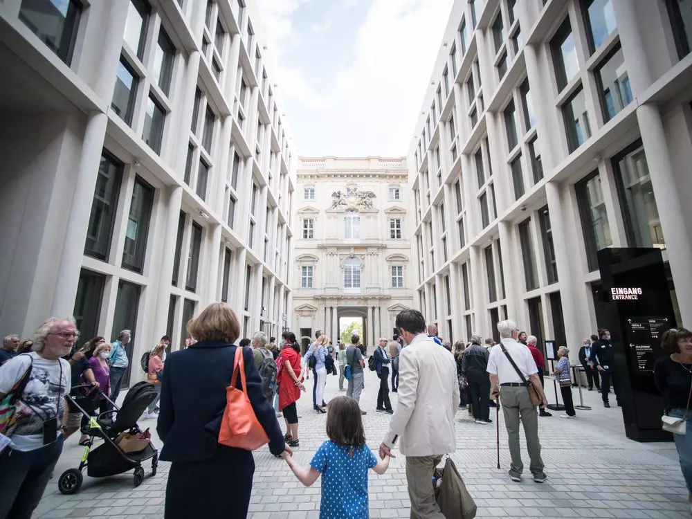 Uma vista da entrada moderna do novo museu, com multidões de pessoas caminhando por um grande pátio cercado por altos pilares brancos e janelas