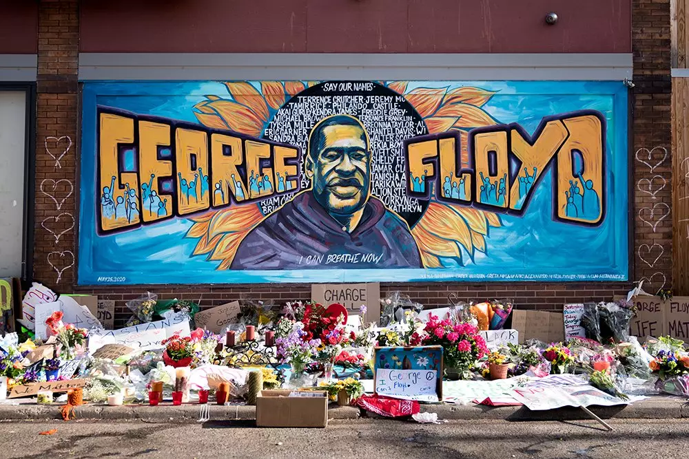 Gran mural en una paret de maó les paraules George Floyd en lletres grogues sobre un fons blau i un retrat d'un home al centre contra un gira-sol amb els noms enumerats amb lletres blanques. Hi ha cors blancs dibuixats al costat del mural i una flor