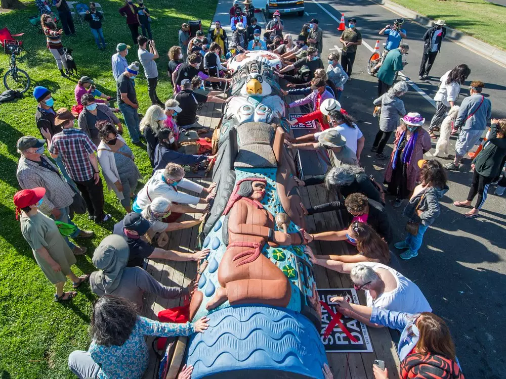 Група людей простягається і торкається стовпа, який лежить боком уздовж кузова вантажівки, пофарбованого в яскраві кольори з фігурою жінки, яка стоїть на колінах.