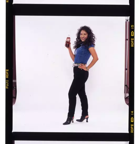 Selena cun chaleco azul e pantalóns negros sostendo unha botella de Coca-Cola