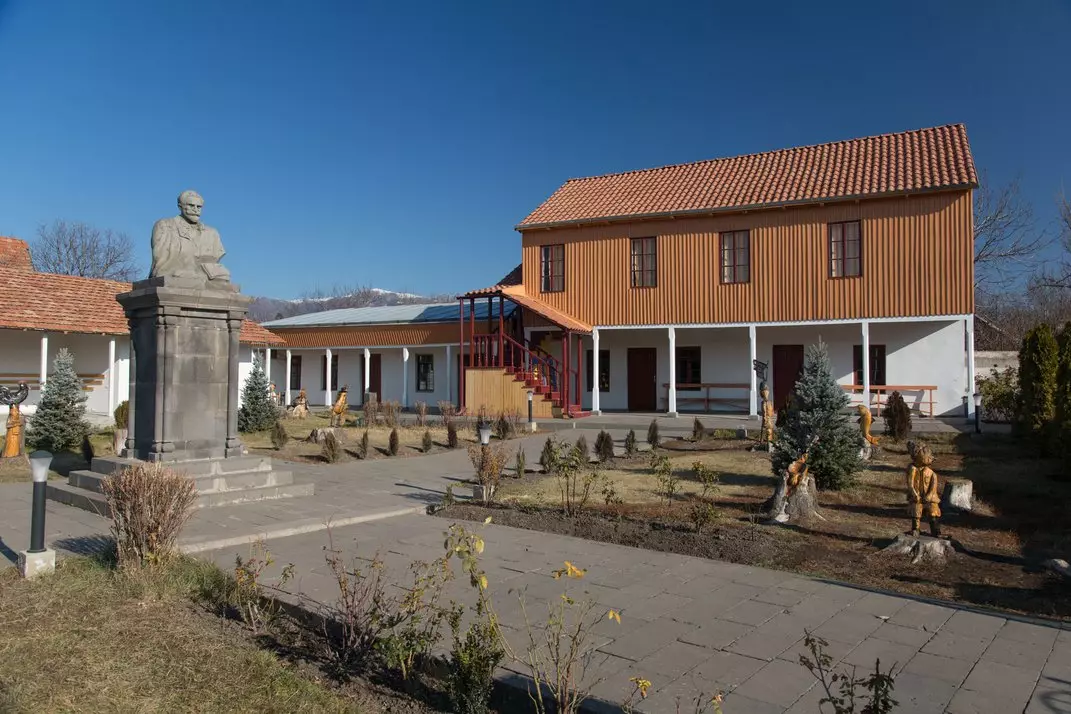 Un patio cun camiño de pedra que conduce a unha escultura central está diante dunha casa atractiva, pintada de branco no primeiro andar e revestida con lamas de madeira estreitas no segundo.