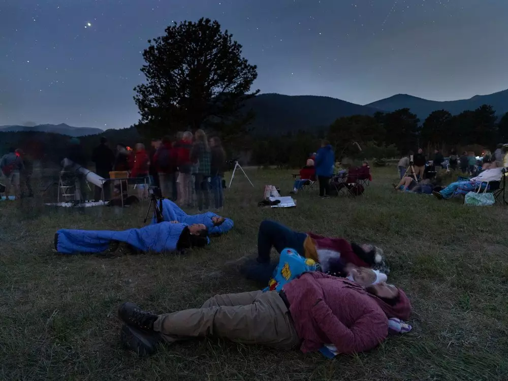 Тълпа от хора лежат на земята в тревисто поле през нощта с планини на заден план