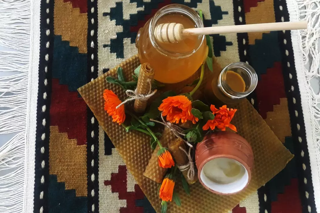 Върху одеяло с геометрични шарки е внимателно подредена мазилка от пчелен восък, мед в буркани, меден крем в теракотена саксия и портокалови цветя.