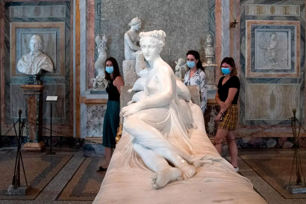 Pohled na sochu v Římě, ležící nohama směrem k fotoaparátu, všechny doplňky nedotčené. Vzadu se na sochu dívají tři turisté v maskách.