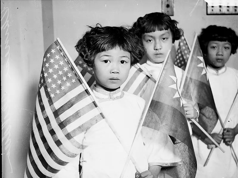 Retrato de grupo de três crianças chinesas em uma sala em Chicago, Illinois, cada uma segurando uma bandeira americana e uma bandeira chinesa, 1929