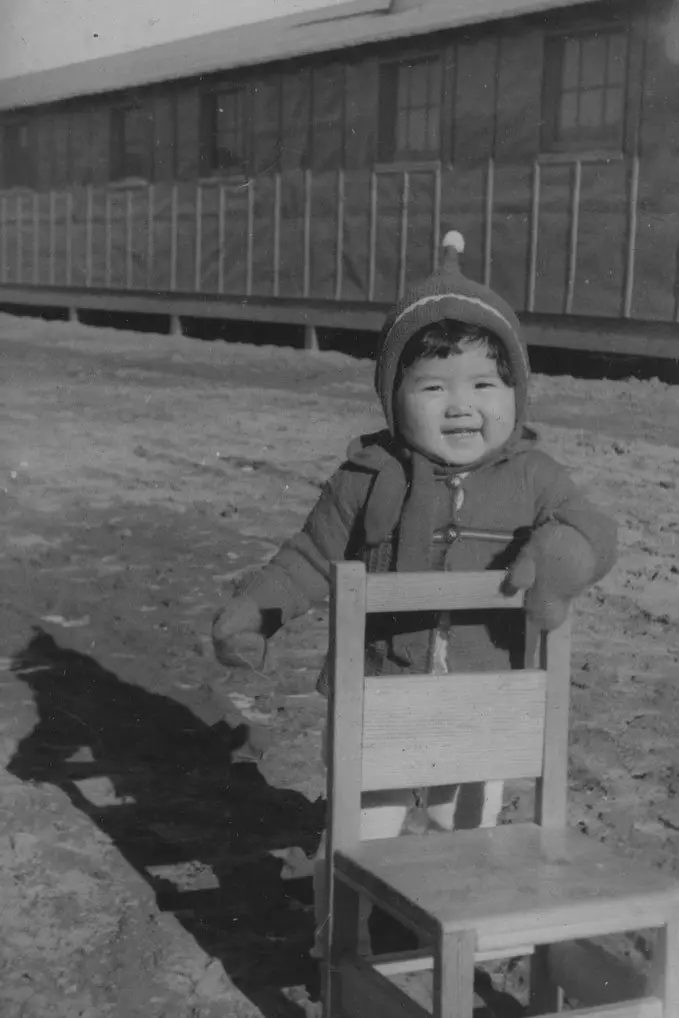 O fotografie alb-negru arată un copil mic zâmbind și ținând un scaun în fața unui gard din zale.