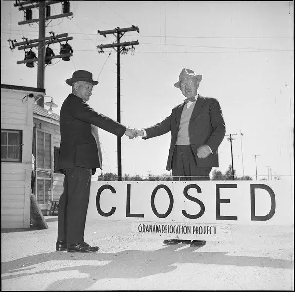 Unha foto en branco e negro mostra a dous homes dándose a man diante dun cartel que di