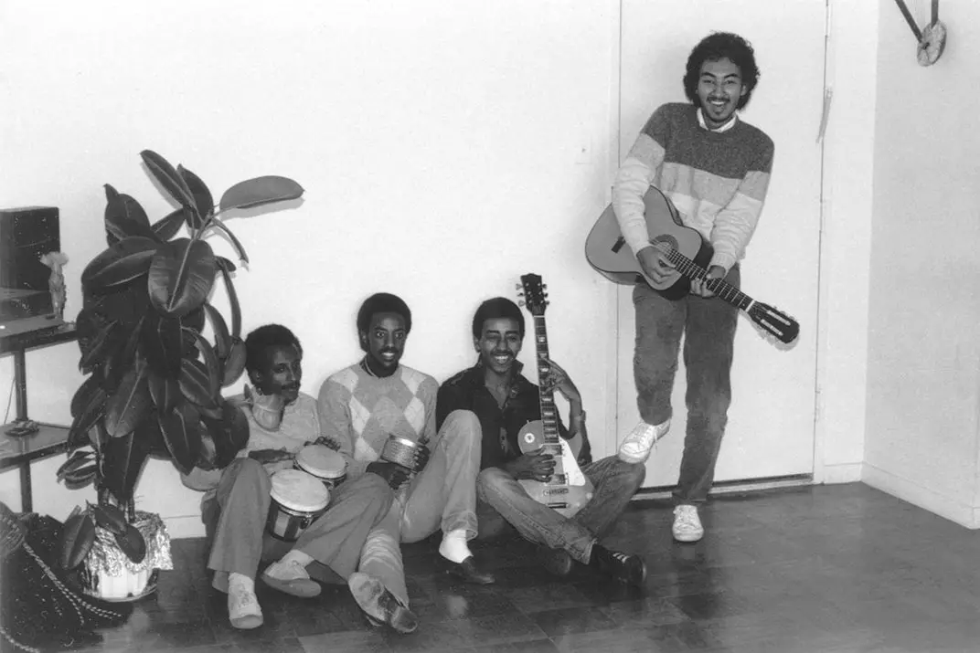 Quatro homens posam, três sentados no chão com uma planta da casa se elevando sobre eles. Todos possuem instrumentos: bongôs, shaker, guitarras elétricas e acústicas. Foto em preto e branco.