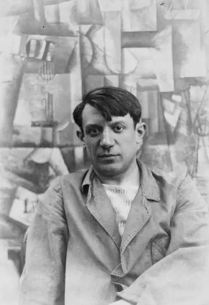 Une image en noir et blanc d'un jeune Picasso assis devant l'un de ses tableaux